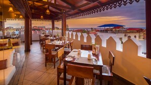 Um restaurante com mesas e cadeiras de madeira, vista para o mar e um pôr do sol ao fundo.