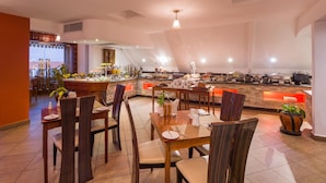 Um espaço de refeições moderno em hotel, com uma ilha central, diversos arranjos de assentos e uma cozinha ao fundo.