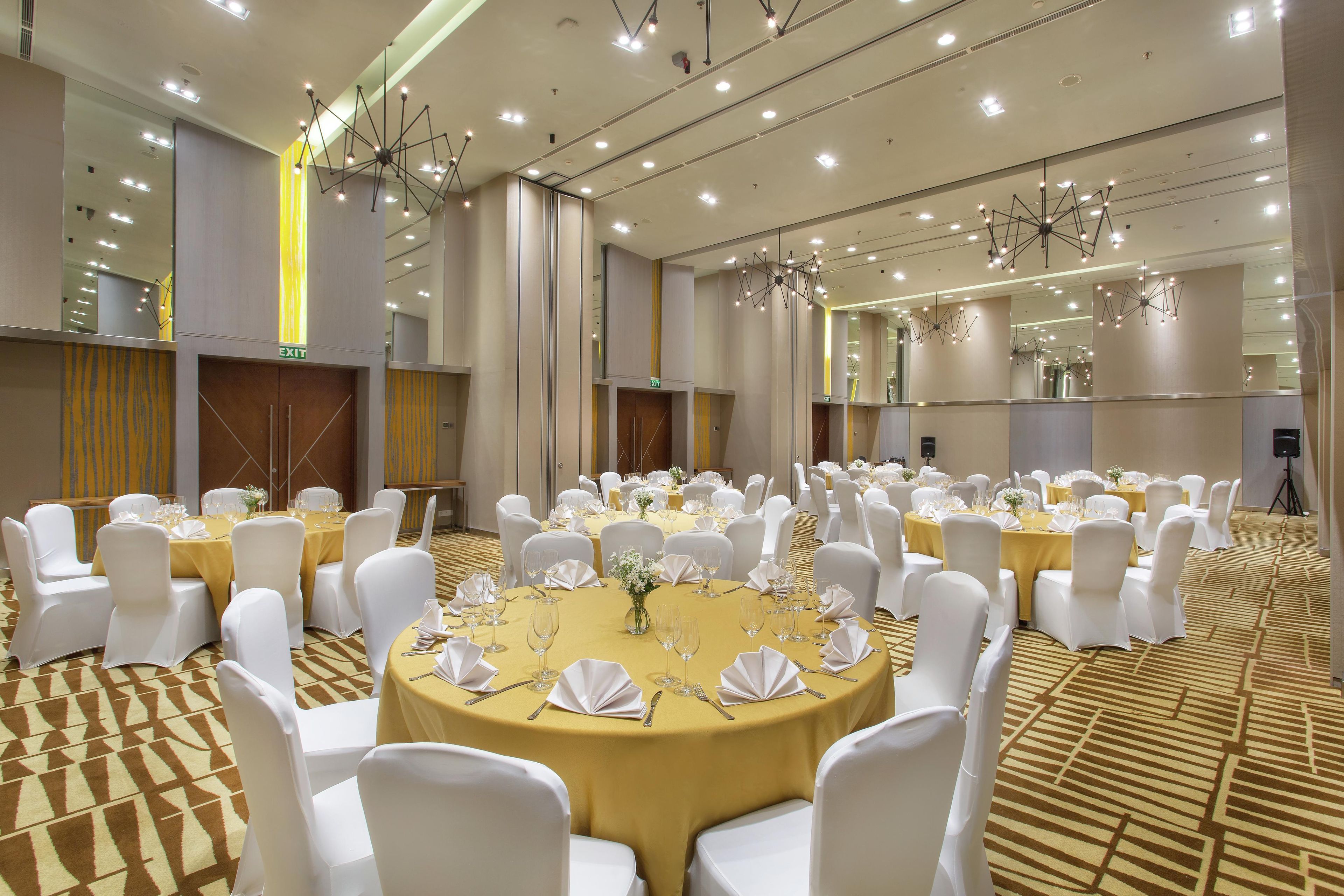Salão de banquetes com mesas redondas, cadeiras brancas e toalhas amarelas.