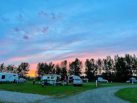 Um camping com veículos recreativos estacionados em um caminho de cascalho, mesas de piquenique e um campo gramado.