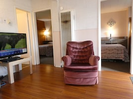 Um quarto com piso de madeira, televisão sobre um suporte, poltrona confortável, cama com colcha estampada e luminária fixada na parede.