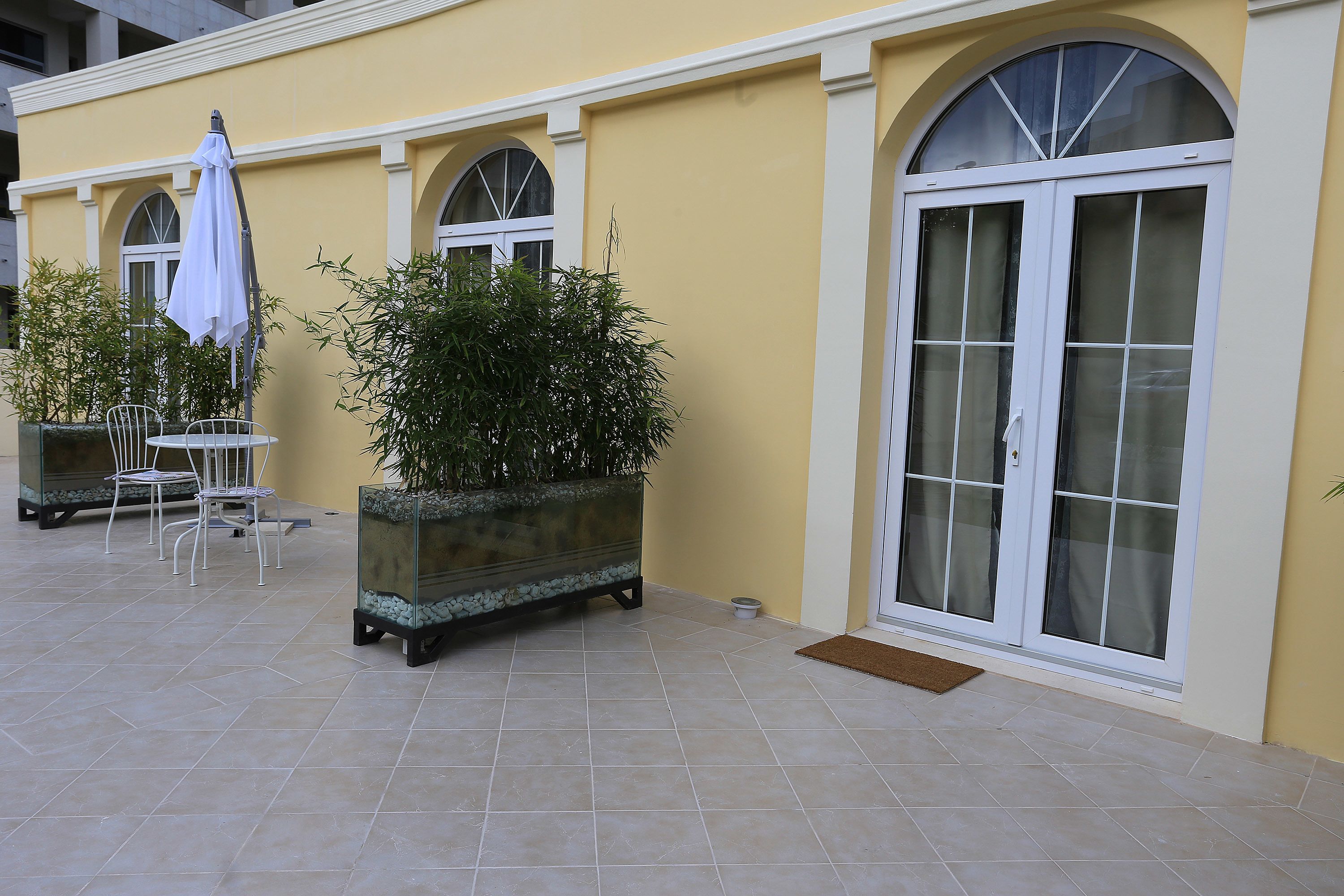 Um edifício amarelo com um pátio ladrilhado, cadeiras brancas, uma mesa e um guarda-sol coberto.