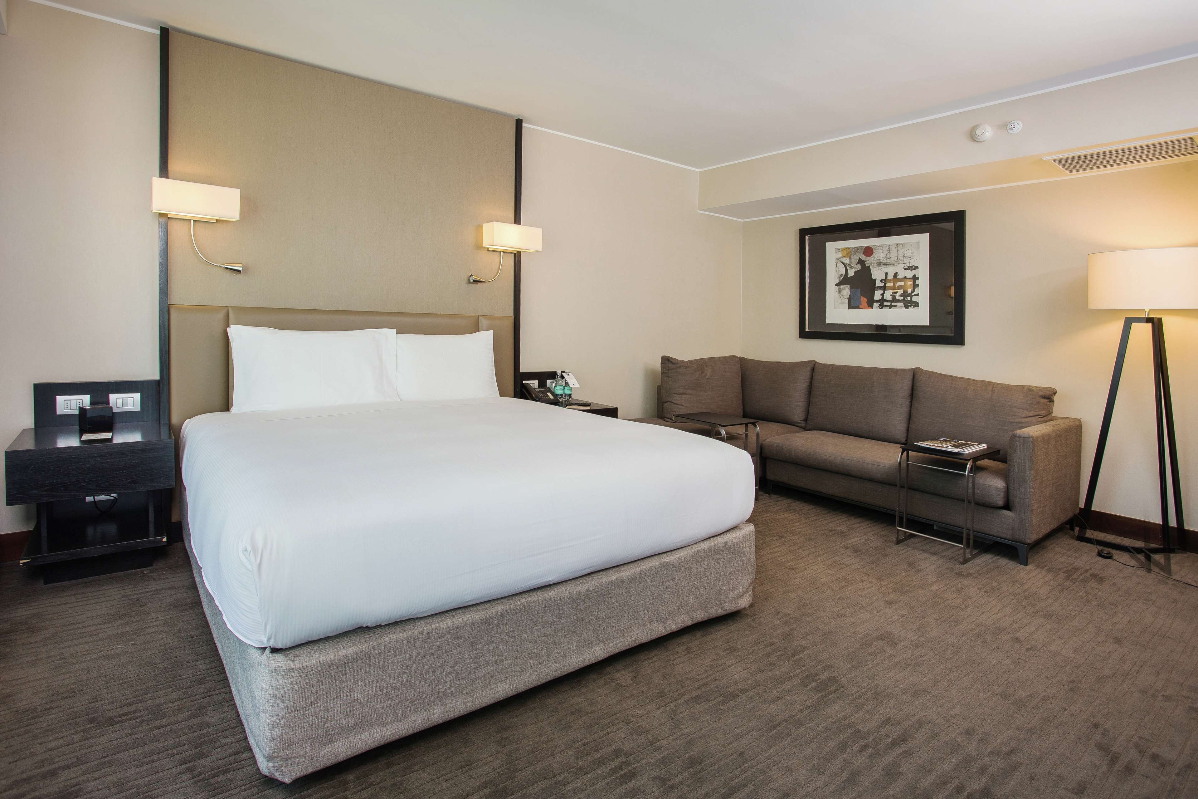 Quarto de hotel com uma cama grande, um sofá, uma escrivaninha e uma luminária.
