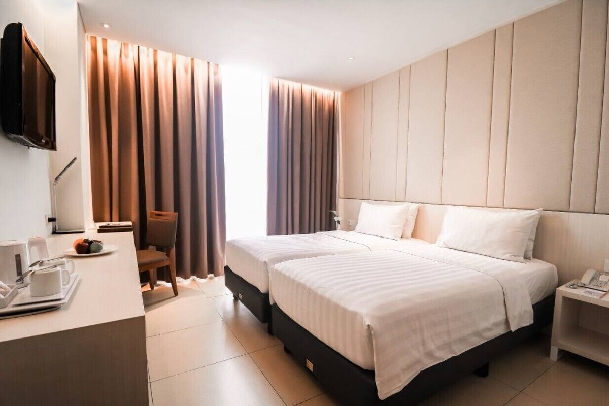 Quarto de hotel com duas camas, uma escrivaninha com telefone e uma chávena, e uma televisão na parede.