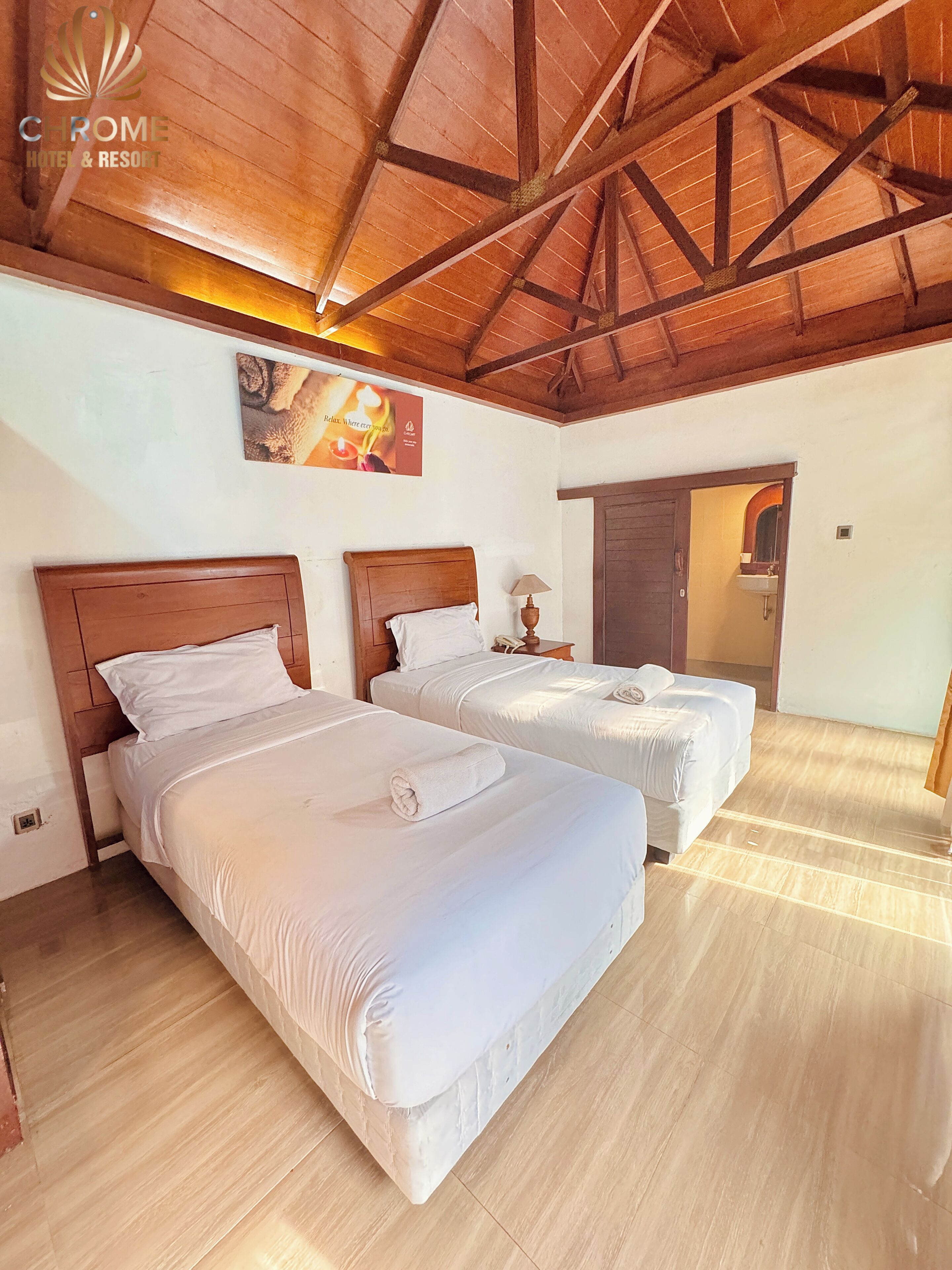 Quarto de hotel com duas camas, vigas de madeira no teto, um quadro pendurado na parede e uma porta que dá para o banheiro.