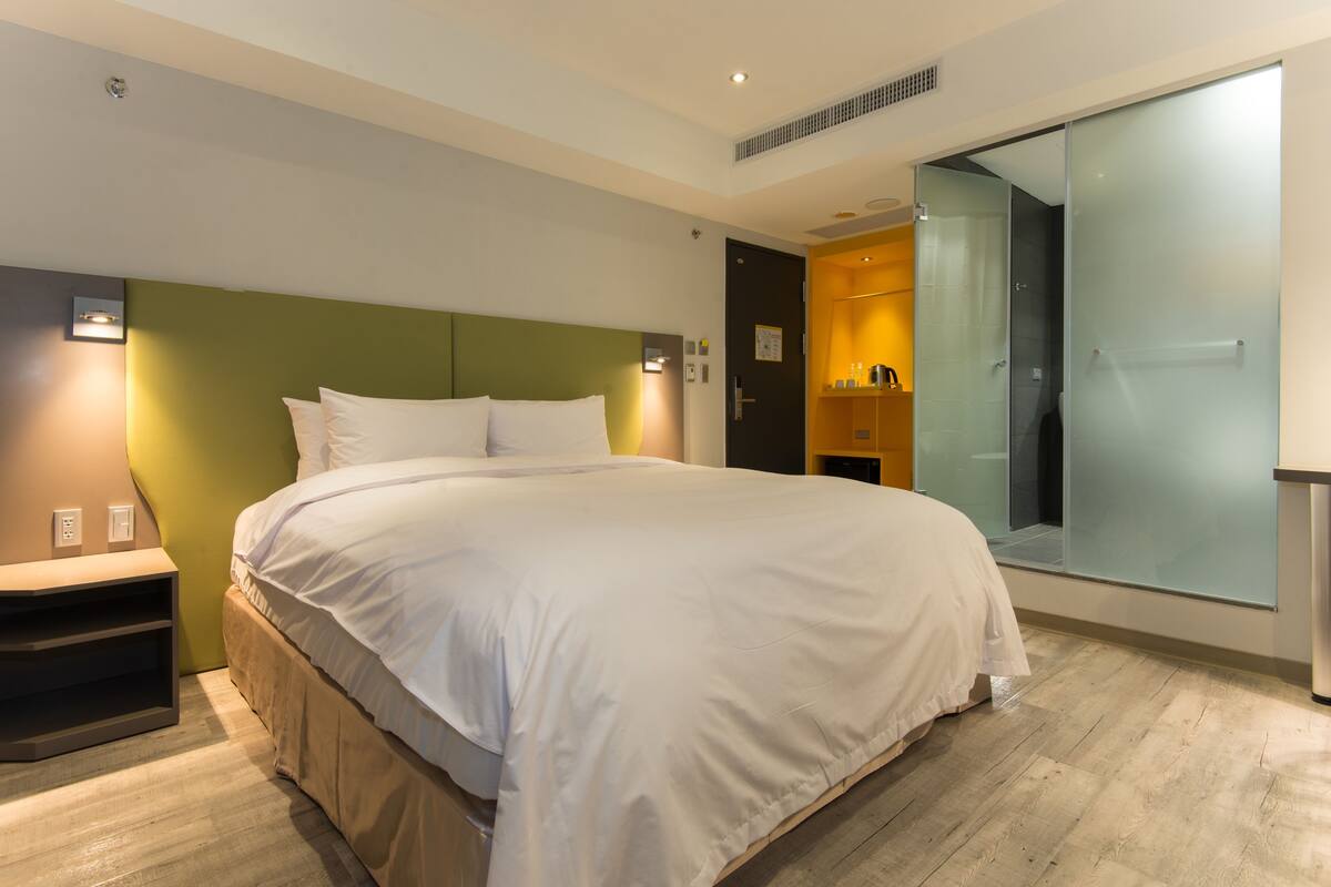 Quarto de hotel moderno com uma cama grande, mesa de cabeceira, chuveiro com paredes de vidro e um pequeno espaço de estar.