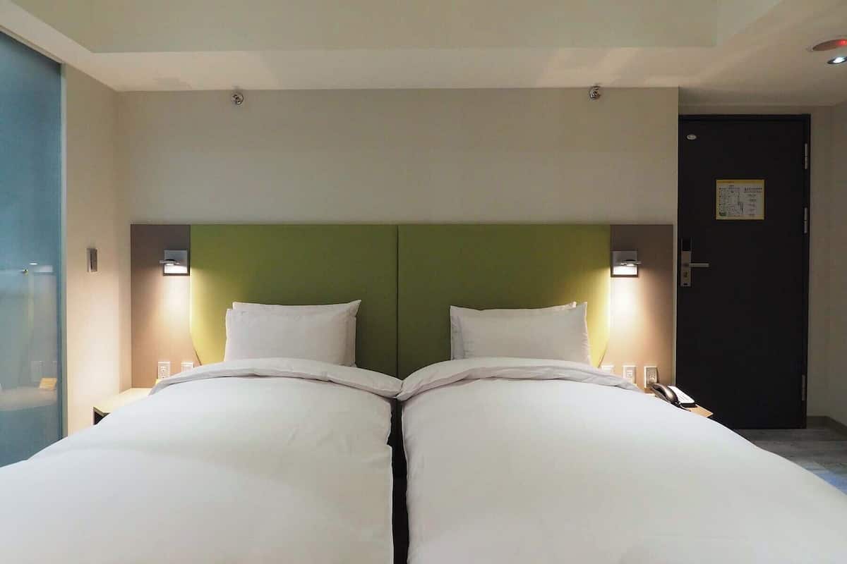 Uma cama de casal com cabeceira verde, duas luminárias de cabeceira e uma porta à direita.