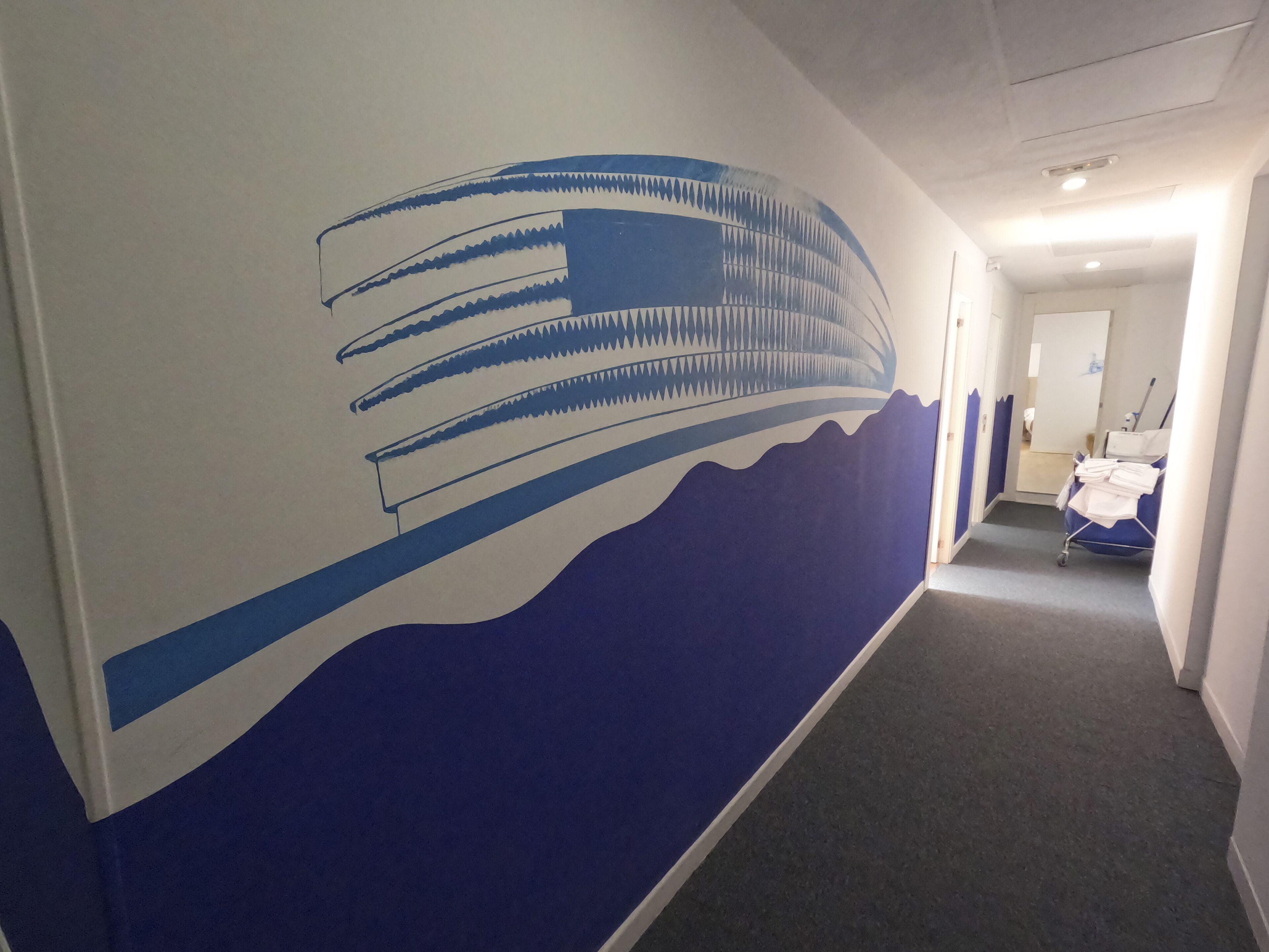 Um corredor com um grande mural azul e branco na parede, piso coberto por carpete, e uma cama com cabeceira azul ao fundo.
