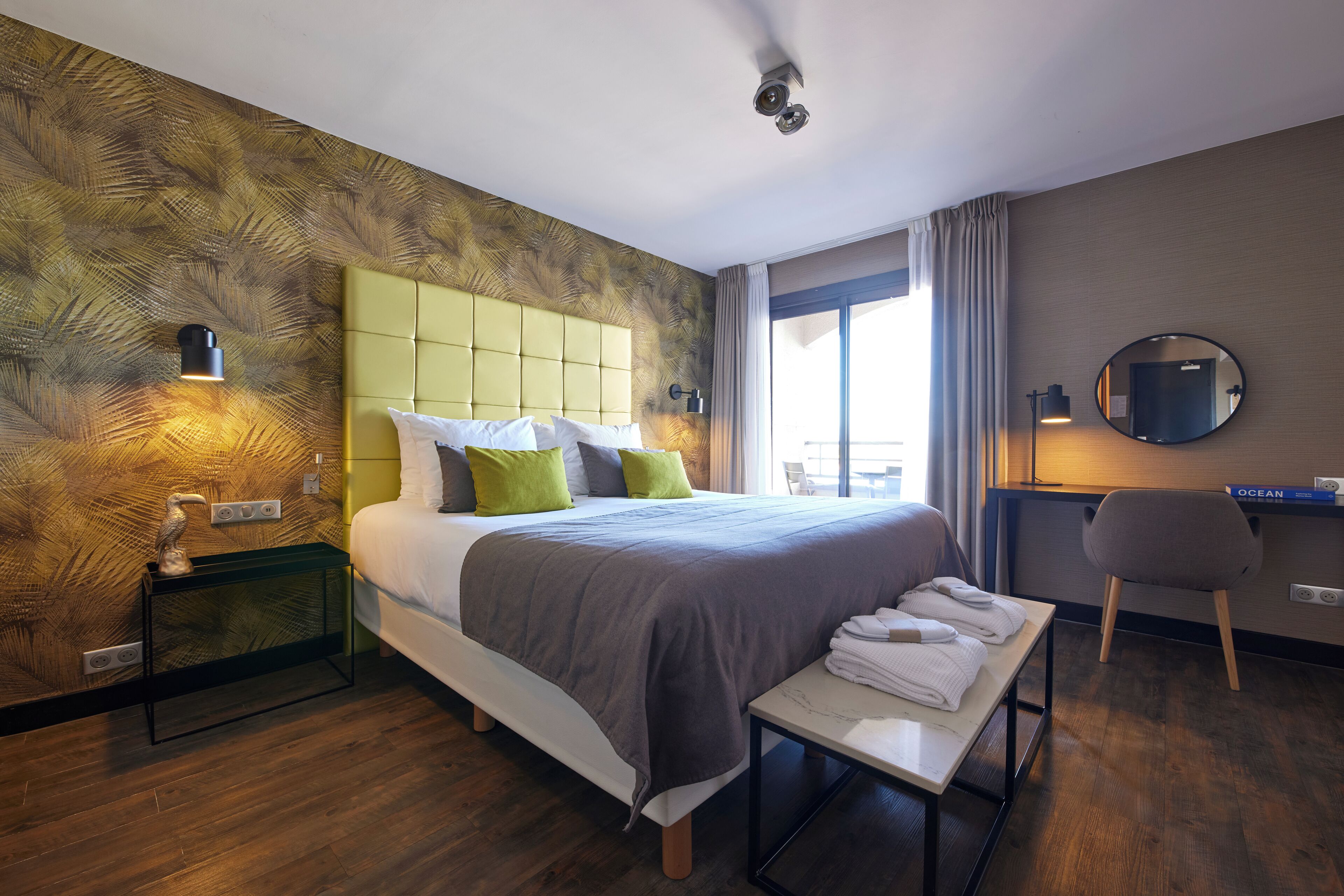 Quarto de hotel moderno com uma cama grande, uma mesinha de cabeceira, uma escrivaninha com cadeira e uma janela ampla.