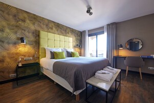 Quarto de hotel moderno com uma cama grande, uma mesinha de cabeceira, uma escrivaninha com cadeira e uma janela ampla.