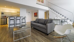 Sala de estar moderna com um sofá cinzento, uma mesa de centro e uma poltrona de balanço. Há uma cozinha com micro-ondas e frigorífico. Uma escadaria leva a um andar superior.