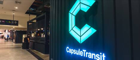 Uma placa da CapsuleTransit com um logotipo geométrico, um carrinho de bagagem e uma pessoa perto de um balcão.
