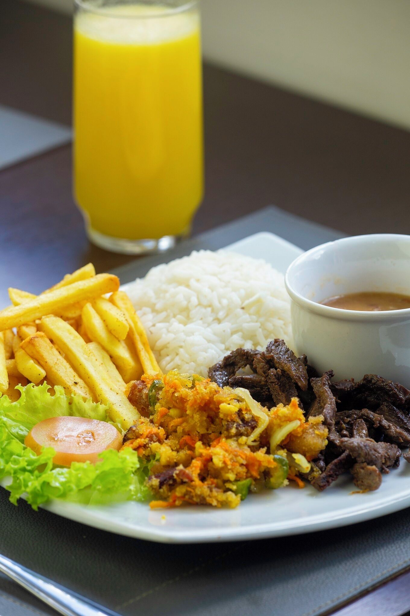 Um prato com arroz, batatas fritas, carne e salada, acompanhado por um copo de suco de laranja e uma pequena tigela de molho.