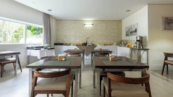 Uma sala de jantar moderna com várias mesas e cadeiras, uma cozinha ao fundo e vista para árvores pela janela.