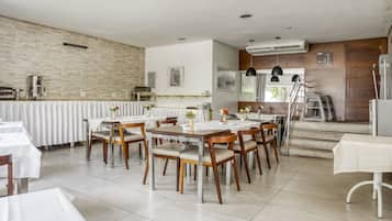 Uma área de refeições moderna com parede de tijolos, mesas e cadeiras brancas. Há uma cozinha com geladeira e ar-condicionado.