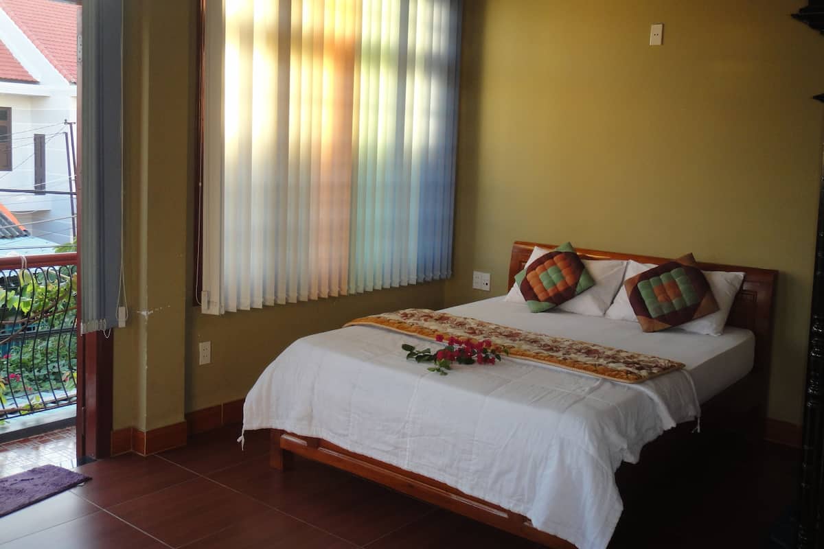 Quarto com cama, janela com persianas, cabeceira de madeira e vista para o exterior.