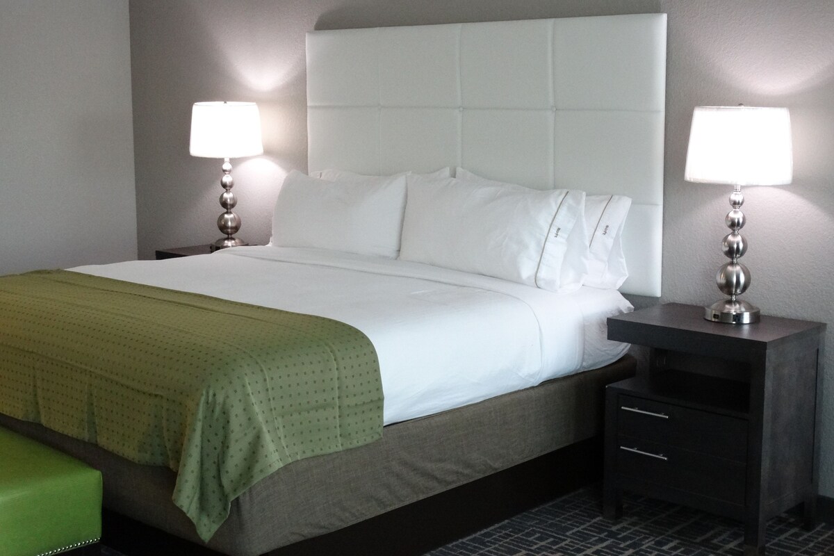 Cama bem arrumada com lençóis brancos e manta verde, acompanhada por duas mesas de cabeceira com luminárias, em um quarto com design simples e clean.