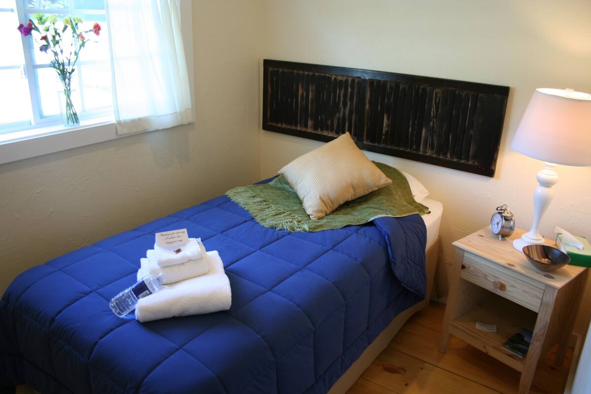 Uma cama bem arrumada com roupa de cama azul, uma mesa de cabeceira com abajur e uma janela com cortinas.