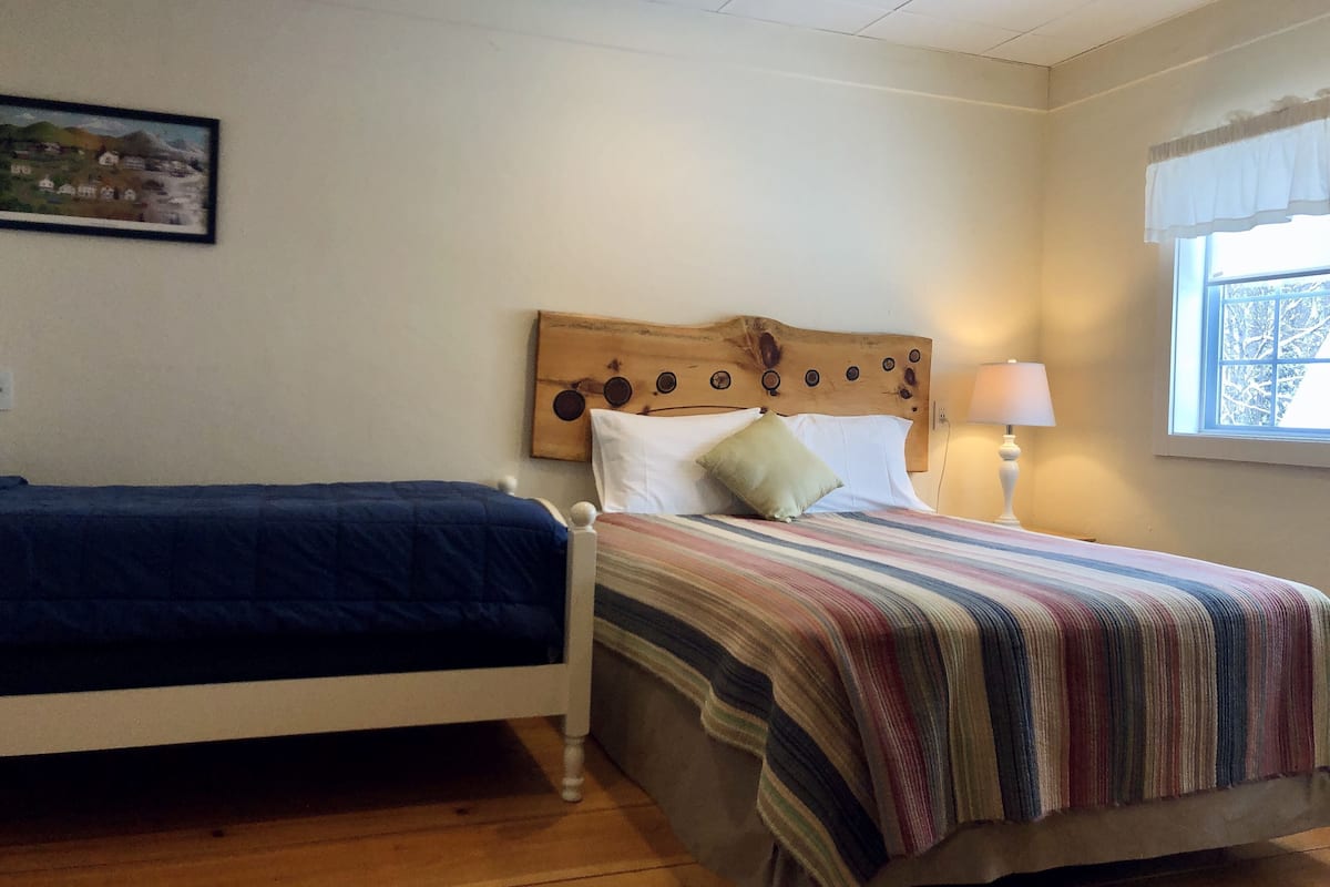 Quarto com duas camas de solteiro, cabeceira de madeira, colcha listrada, luminária de cabeceira e janela com vista para árvores.