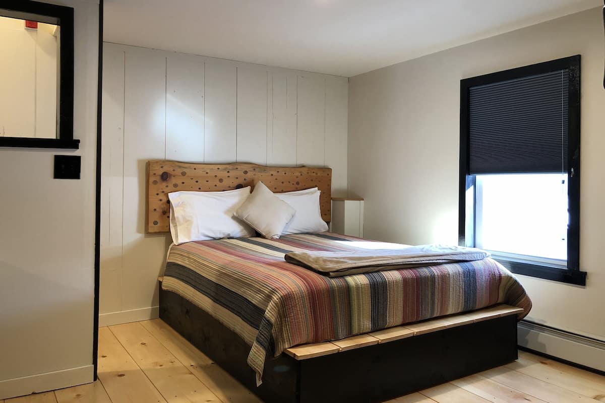 Quarto com cabeceira de madeira, colcha listrada e janela com persianas.