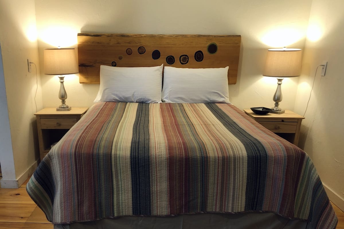 Uma cama com colcha listrada, duas almofadas brancas e cabeceira de madeira com detalhes circulares. Há duas mesas de cabeceira com luminárias de cada lado da cama.