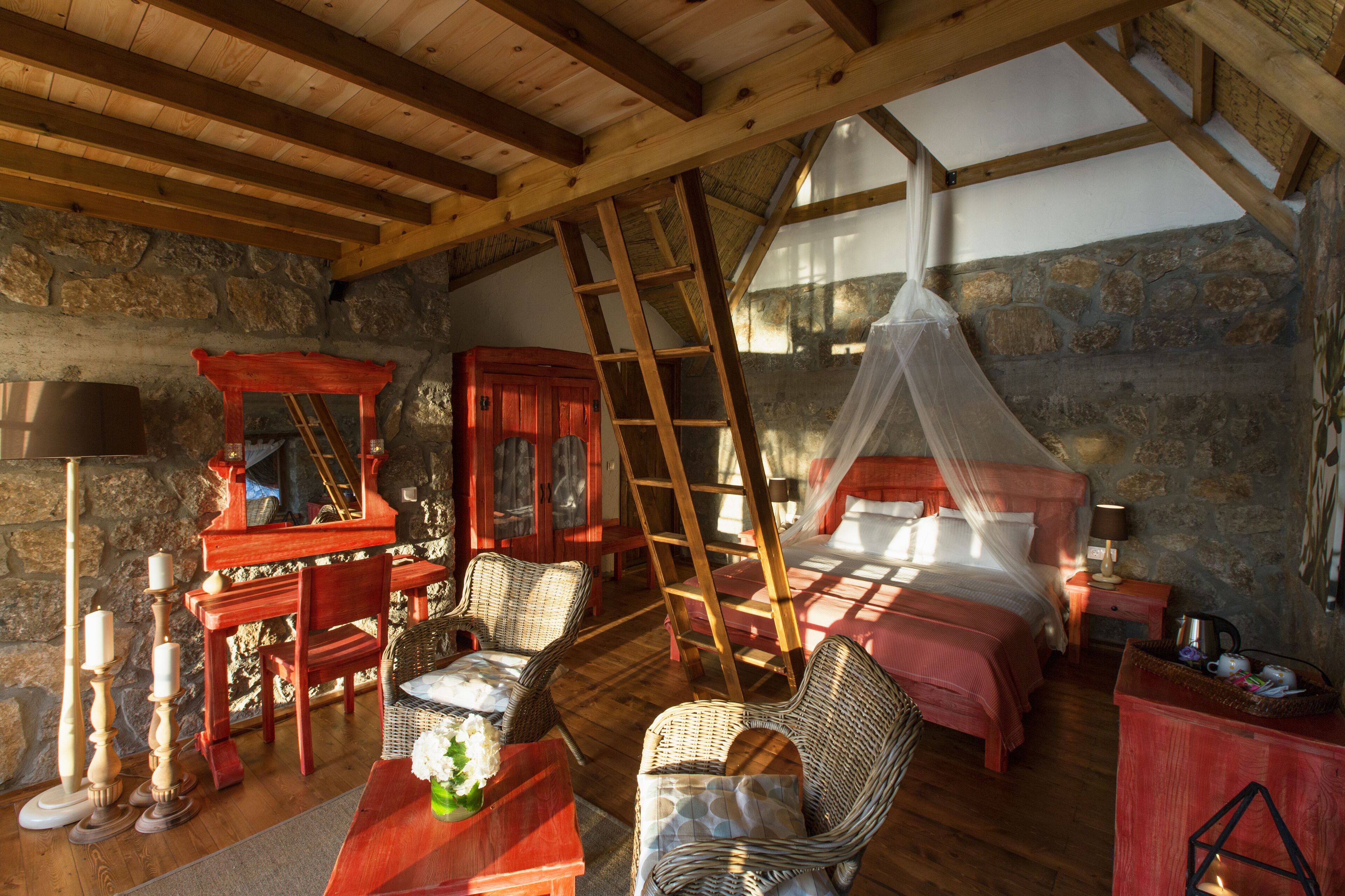 Quarto rústico com parede de pedra, vigas de madeira, cama com dossel, móveis de vime e uma escada.