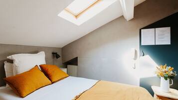 Quarto de hotel moderno com claraboia, cama com roupa de cama branca e laranja, mesa de cabeceira com um vaso de flores e telefone fixado na parede.