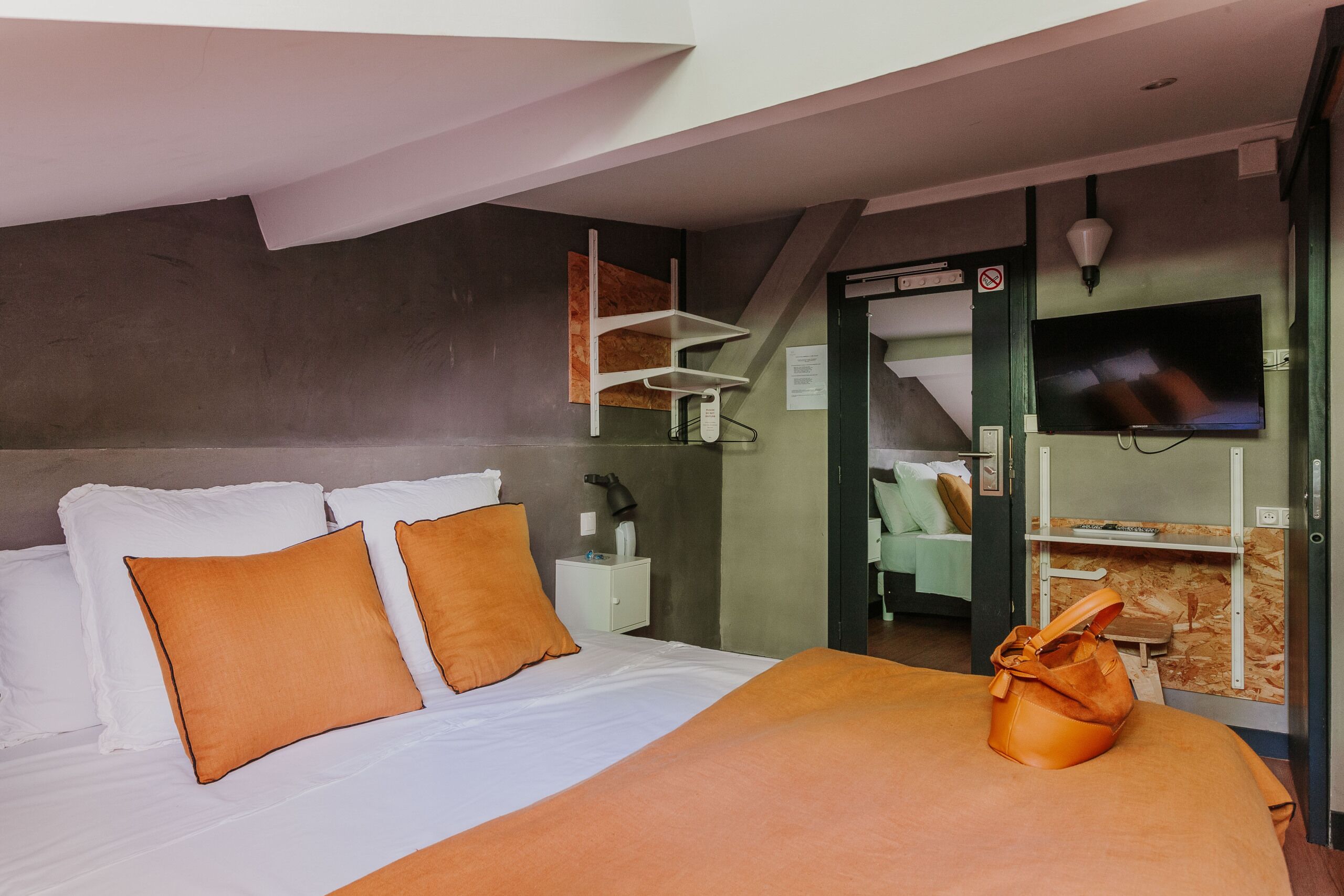 Quarto de hotel moderno com cama, almofadas, mesa de cabeceira, abajur, televisão e sapateira.
