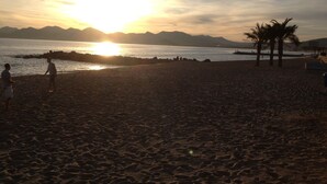 Uma praia com pessoas caminhando e brincando, um pôr do sol ao fundo e palmeiras.