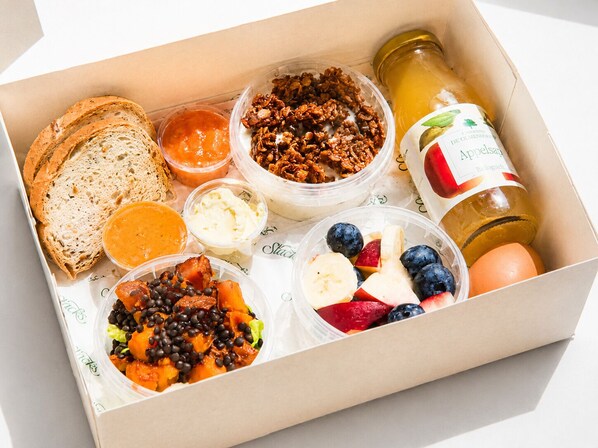 Uma caixa com diversos alimentos, incluindo pão, granola, iogurte e um frasco de molho.