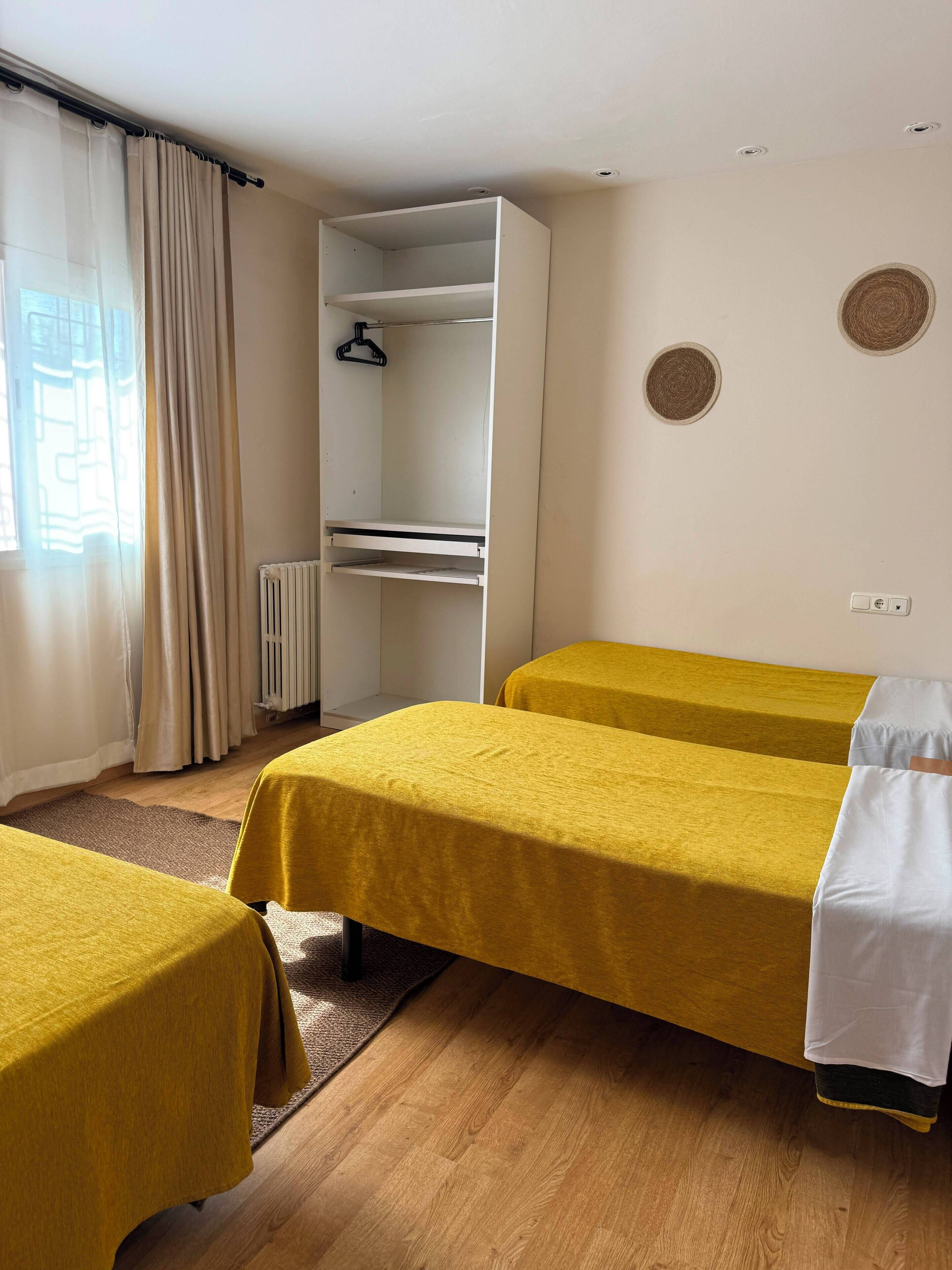 Quarto com duas camas cobertas por lençóis amarelos, um guarda-roupa branco, um radiador e uma janela com cortinas.