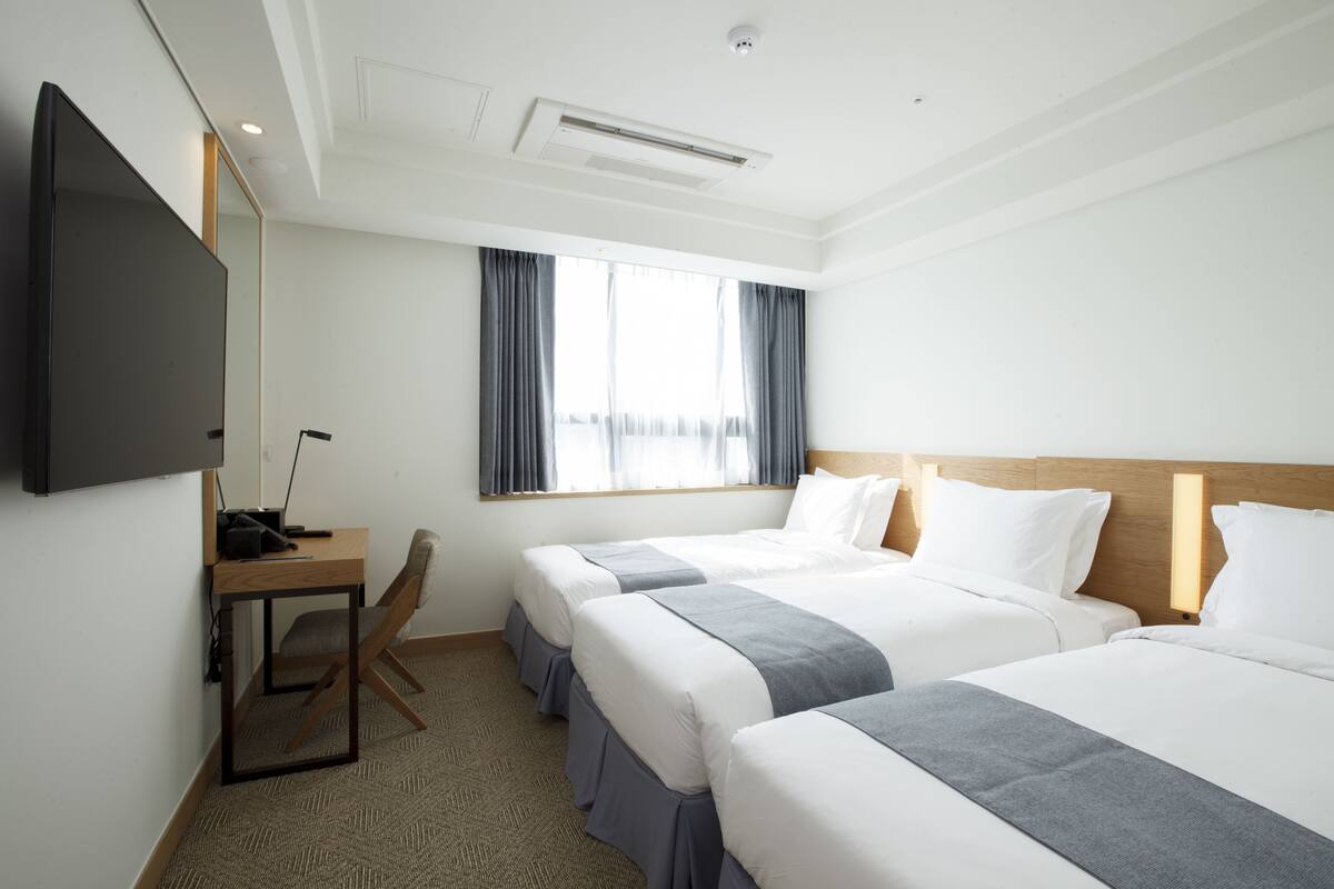 Quarto de hotel com duas camas, uma mesa com cadeira, televisão de tela plana e uma janela grande com cortinas.
