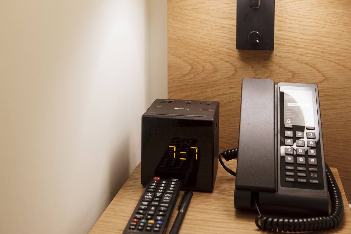 Quarto de hotel com uma mesa de cabeceira contendo um telefone, um controle remoto e uma luminária.