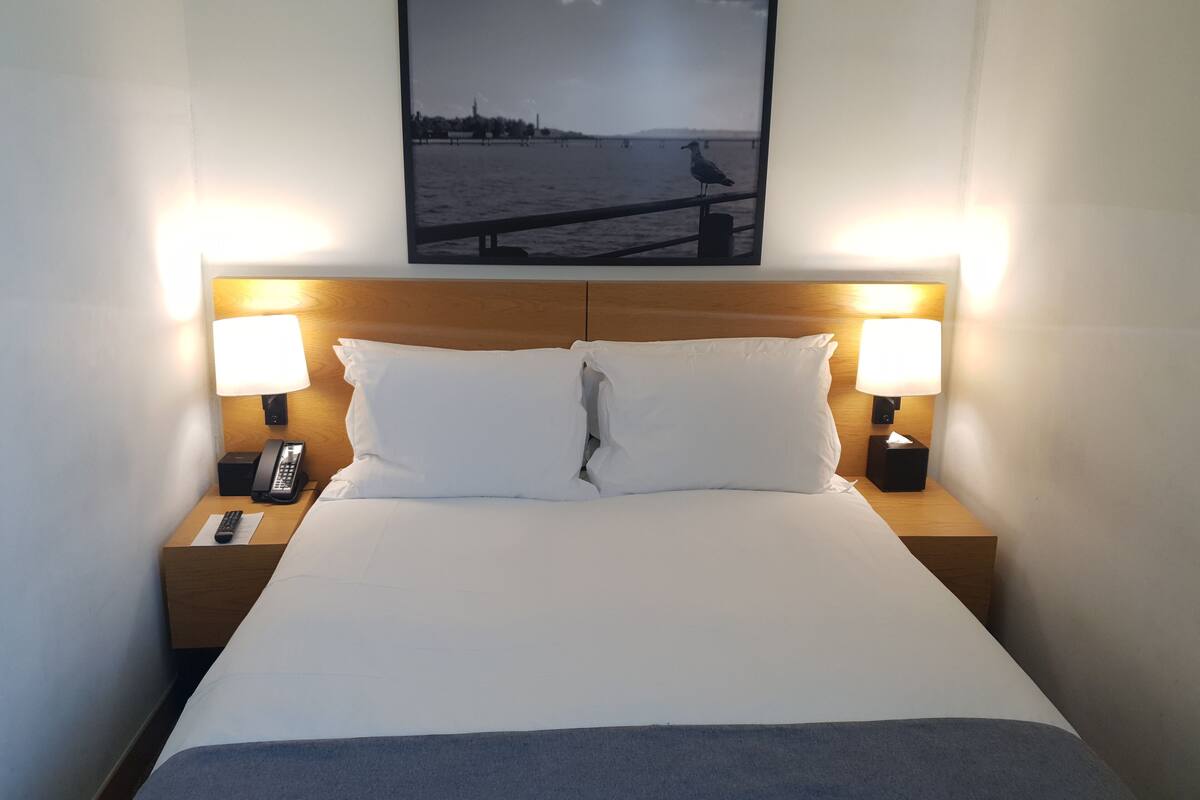 Quarto de hotel com uma cama, duas luminárias de cabeceira, um quadro na parede e um telefone na mesinha de cabeceira.