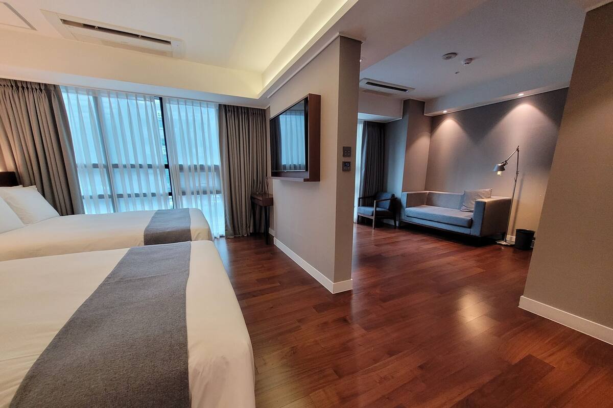 Quarto de hotel moderno com duas camas, um sofá, uma TV e uma janela grande com cortinas.