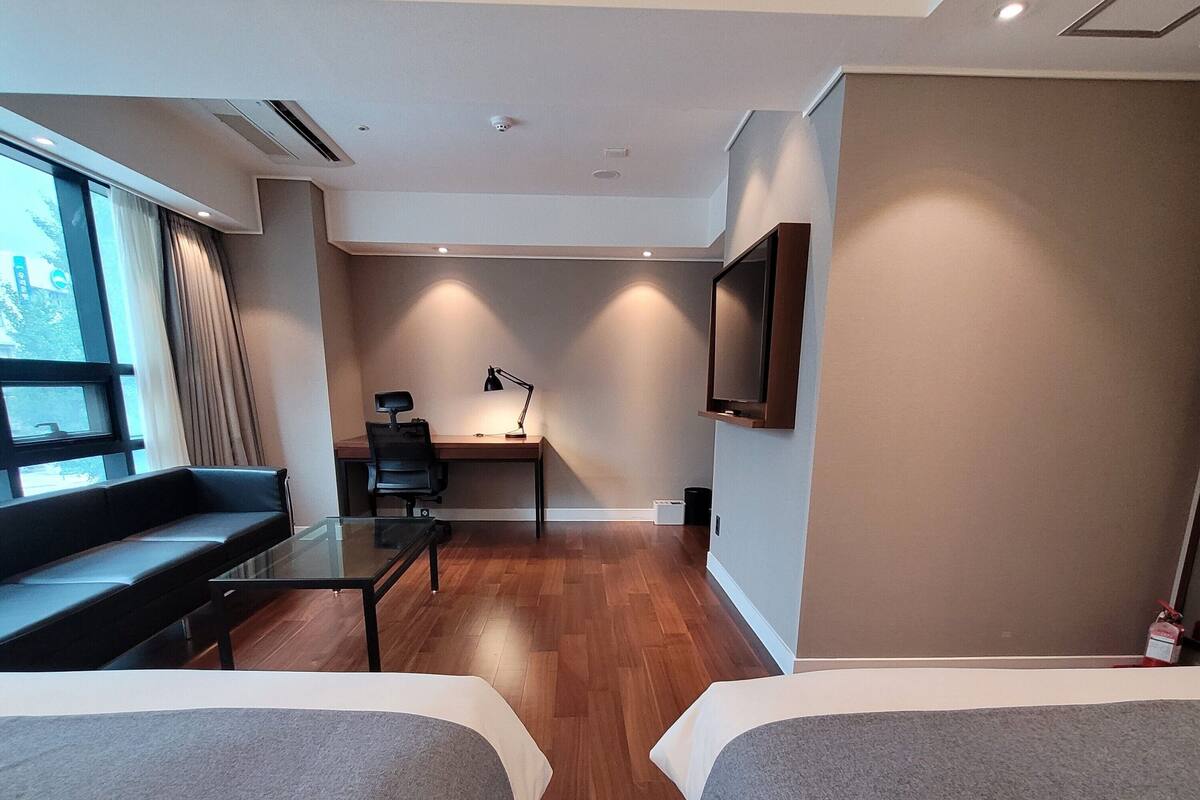 Quarto de hotel moderno com duas camas, uma escrivaninha, uma cadeira, televisão e janela com cortinas.