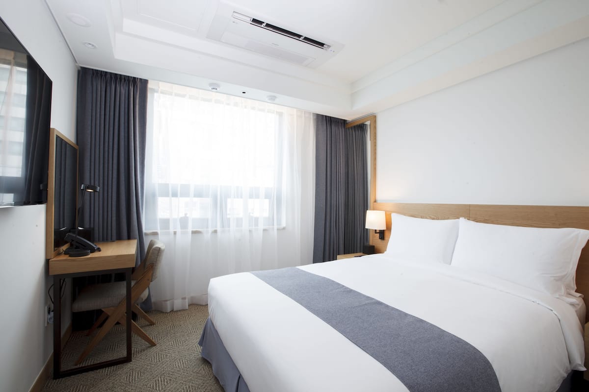 Quarto de hotel com cama, escrivaninha e cadeira, luminária de mesa e janela com cortinas.