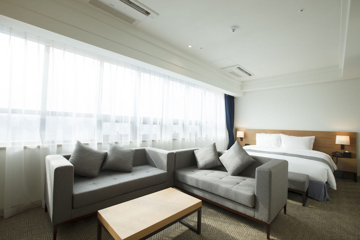 Quarto de hotel com sofá cinza, mesa de centro de madeira, cama com roupa de cama branca e mesa de cabeceira com abajur.