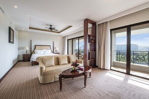 Quarto de hotel com cama, sofá, mesa de centro, estante e varanda com vista.