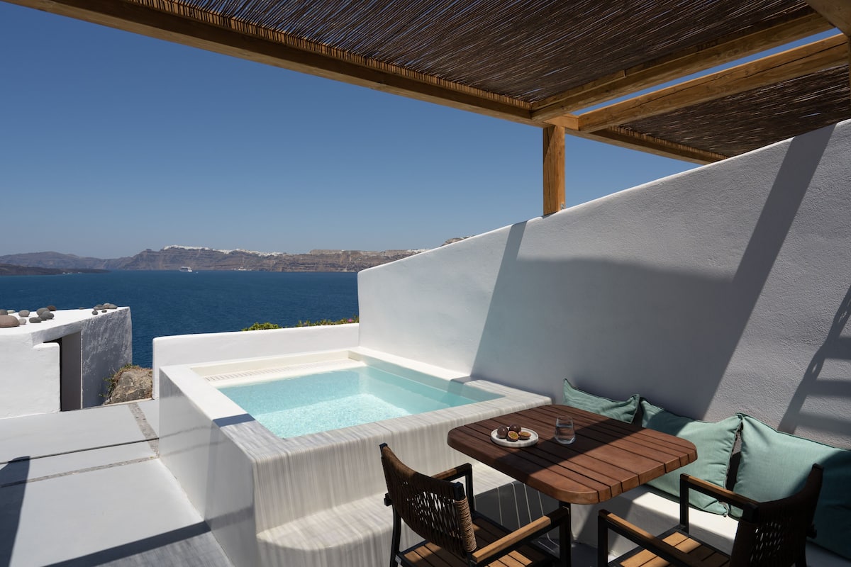 Área de estar ao ar livre iluminada pelo sol, vista para o mar, banheira de hidromassagem e mesa com um prato de comida.
