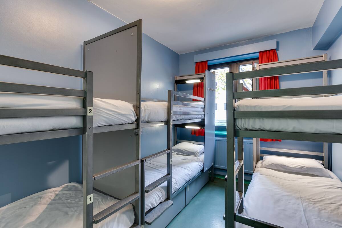 Um dormitório com beliches, camas numeradas e uma janela com cortinas vermelhas.