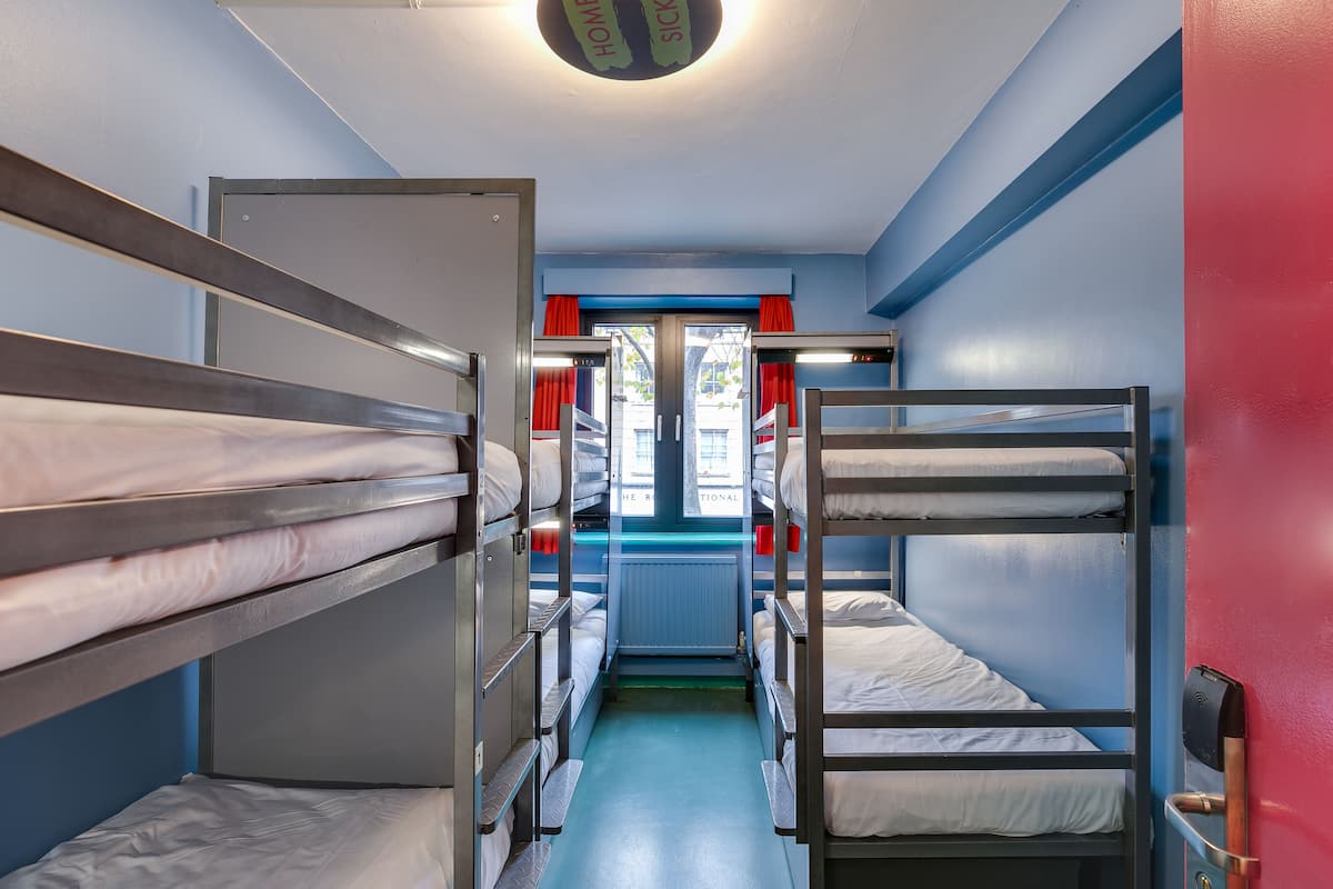 Um corredor estreito com beliches em ambos os lados, uma porta no fim e uma janela com cortinas vermelhas.