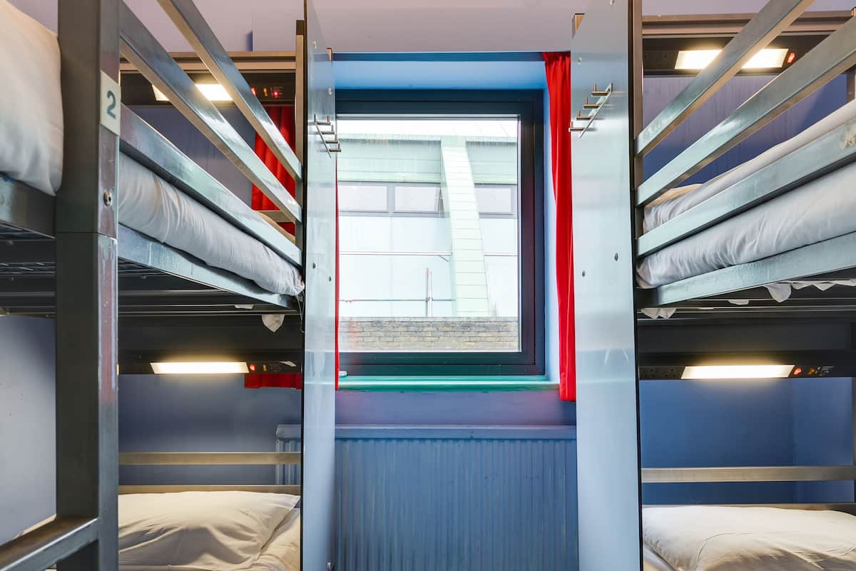 Um vagão de trem com beliches, uma janela com cortinas vermelhas e uma parede azul.