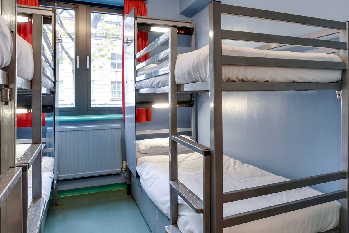 Um dormitório com beliches de metal, uma janela com cortinas vermelhas e um radiador.