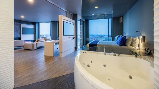 Quarto de hotel moderno com uma banheira grande, um conjunto de sofás, televisão e vista para a cidade.
