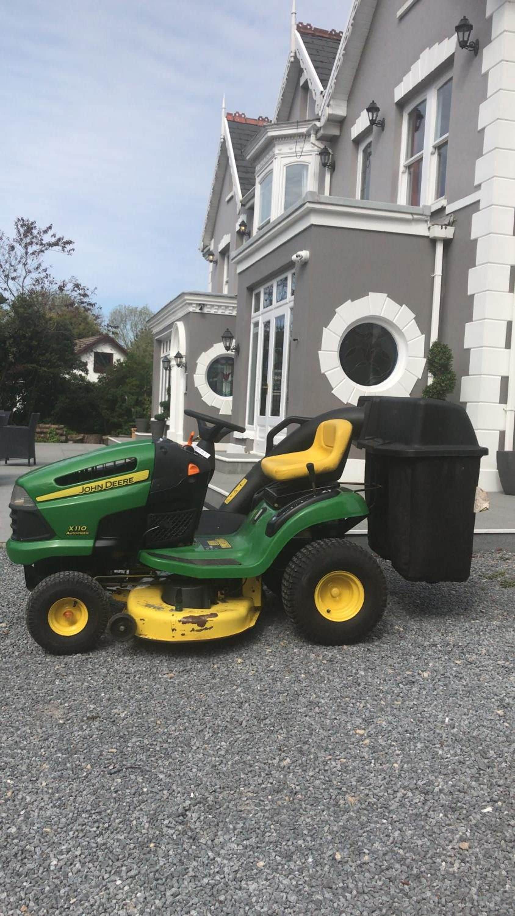 Um cortador de relva verde John Deere X110 com um contentor de lixo preto preso, estacionado em cascalho em frente a uma casa cinzenta com detalhes brancos e janelas circulares decorativas.