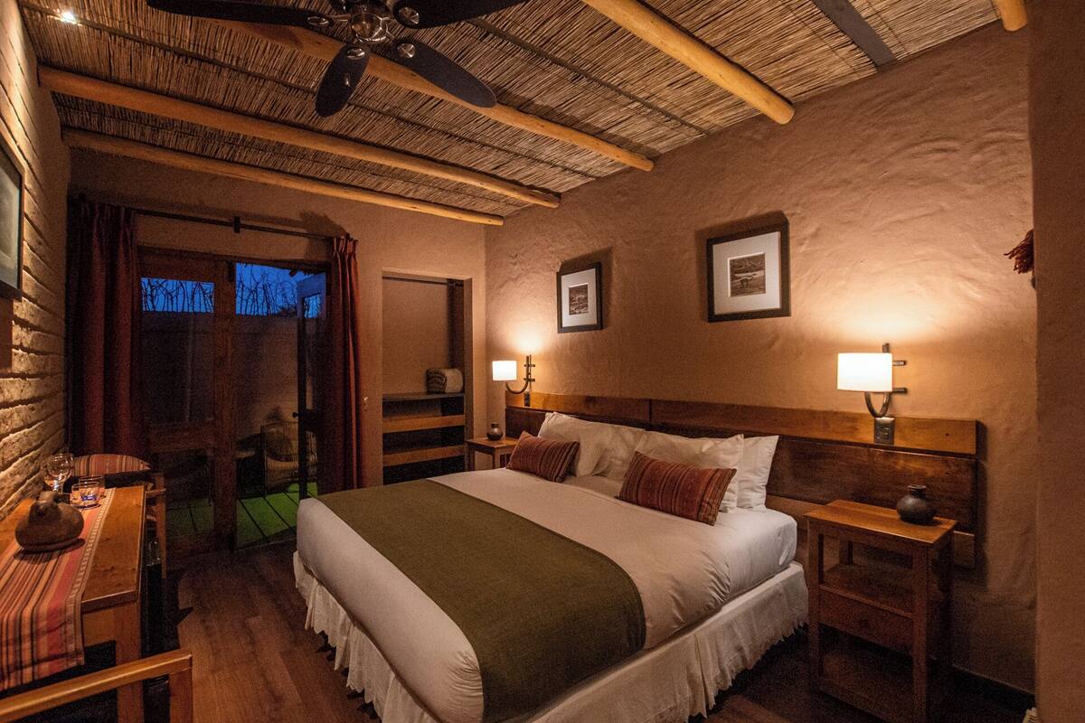 Quarto de hotel acolhedor com uma cama grande, cabeceira de madeira e um ventilador de teto. Há duas mesas de cabeceira com luminárias e uma pequena mesa com um vaso.