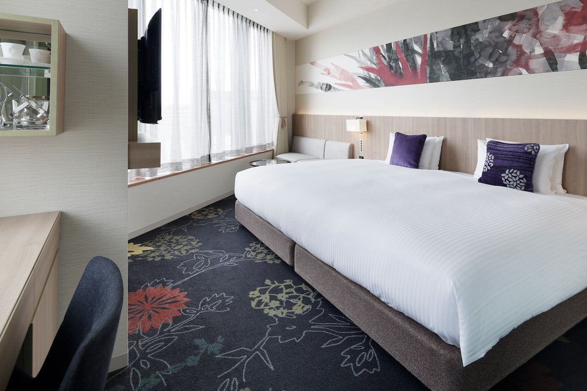 Quarto de hotel com cama, escrivaninha, cadeira, televisão, janela com cortinas, um quadro na parede e luminária.
