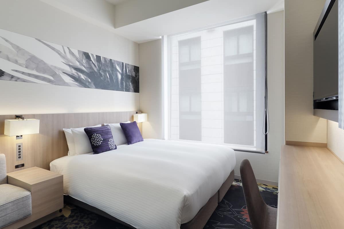 Quarto de hotel com uma cama grande, abajures, mesa de cabeceira, uma cadeira, uma janela ampla e televisão na parede.