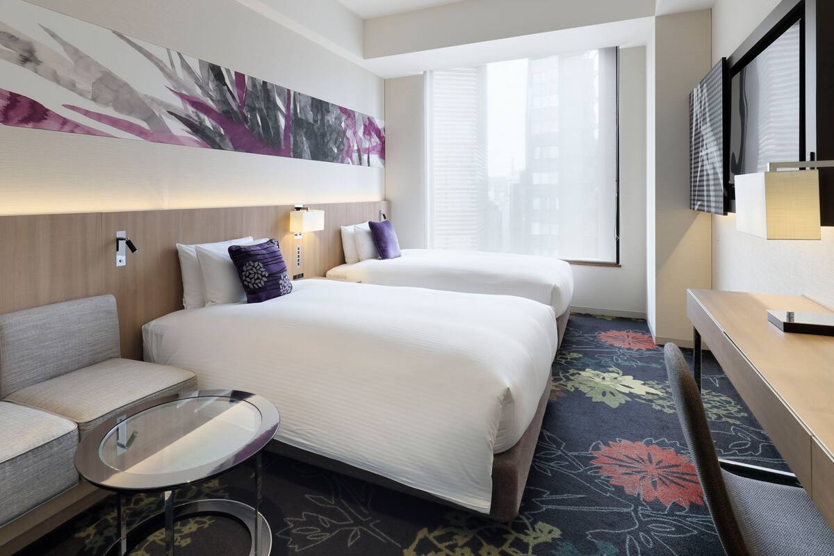 Quarto de hotel com duas camas, um sofá, uma mesinha, uma televisão e uma janela grande.